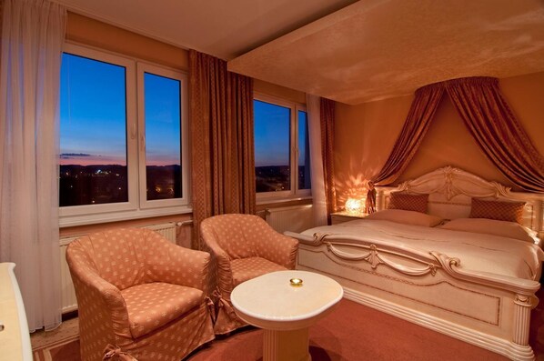 Quarto de hotel com uma cama grande, duas poltronas, uma mesinha e vista da cidade ao entardecer.