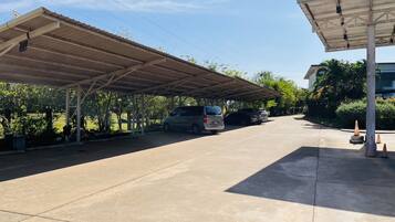 Uma área de estacionamento coberta com veículos, piso de concreto e um prédio ao fundo.