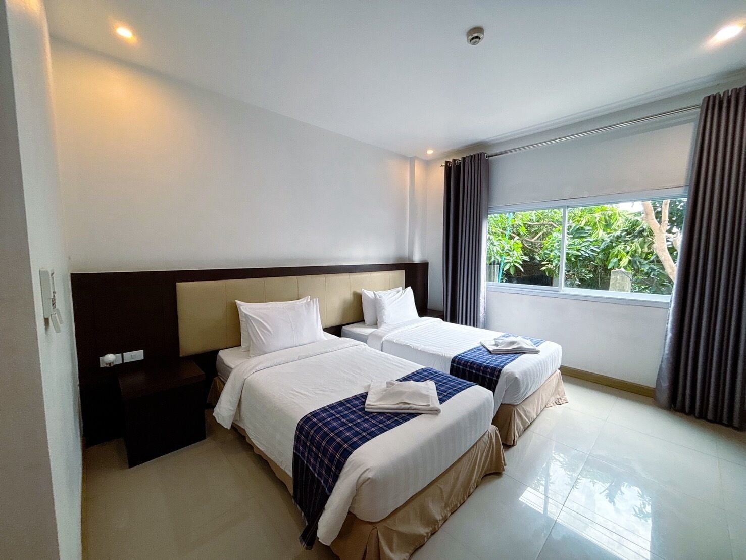 Quarto de hotel com duas camas, uma mesa de cabeceira, janela com cortinas e vista para o verde lá fora.