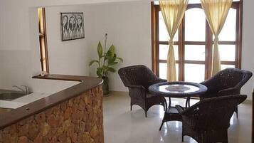 Quarto com bancada de pedra, planta em vaso, cadeiras de vime em torno de uma mesinha e uma janela com cortinas.