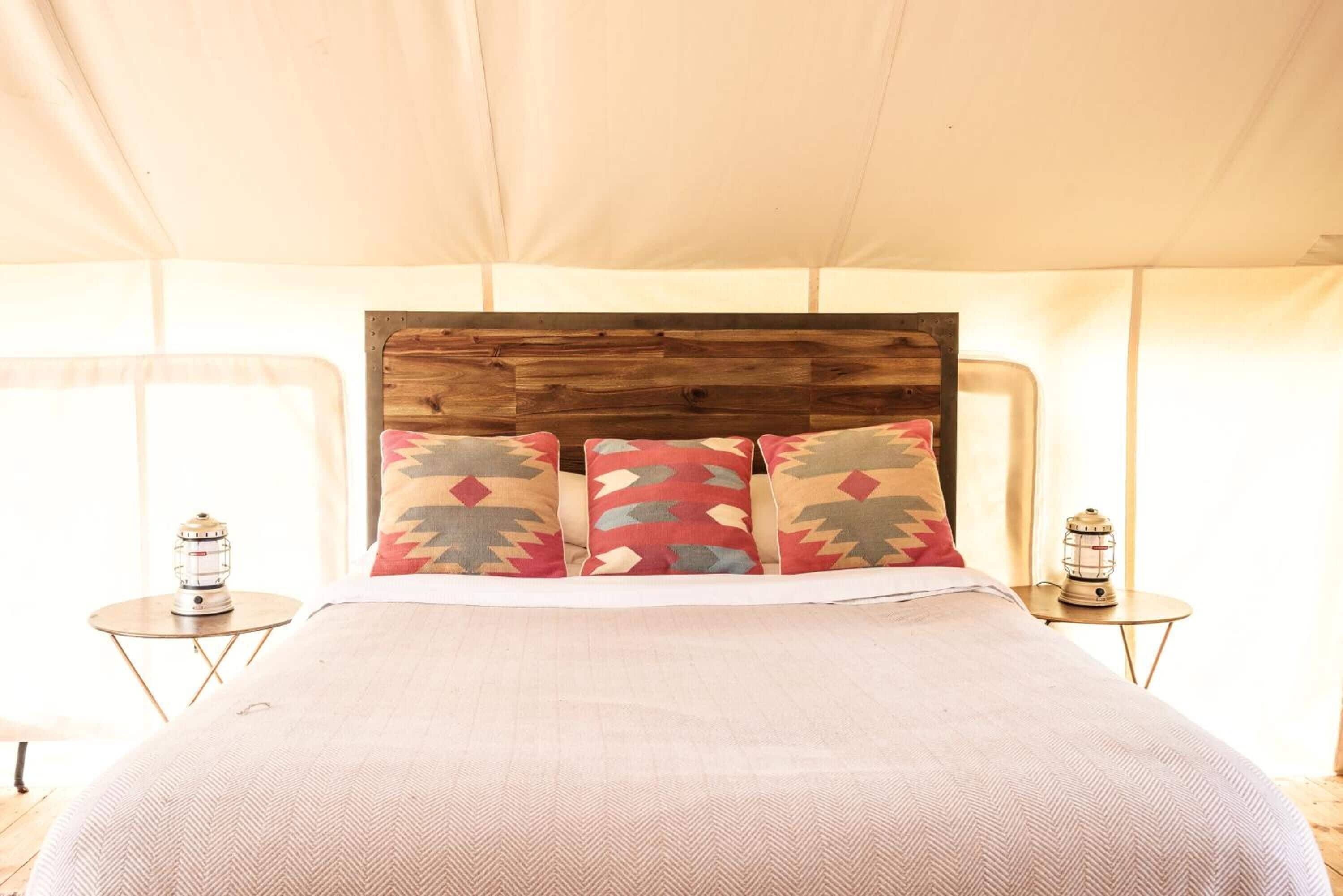 Cama com cabeceira de madeira, três almofadas estampadas e colcha em tom claro. Há duas mesinhas com lanternas de cada lado da cama.