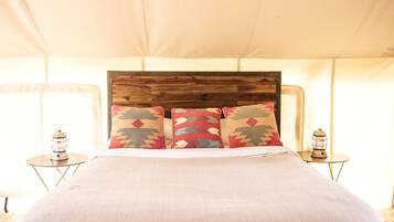 Cama com cabeceira de madeira, três almofadas estampadas e colcha em tom claro. Há duas mesinhas com lanternas de cada lado da cama.