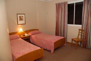 Quarto de hotel com duas camas de solteiro, mesa de cabeceira com luminária, uma cadeira e janela com cortinas.