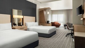 Quarto de hotel com duas camas, uma escrivaninha, uma cadeira, televisão e uma janela grande com cortinas.