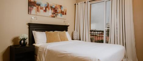 Cama bem arrumada com lençóis brancos e duas almofadas, mesa de cabeceira com um vaso de flores e um grande quadro abstrato na parede.