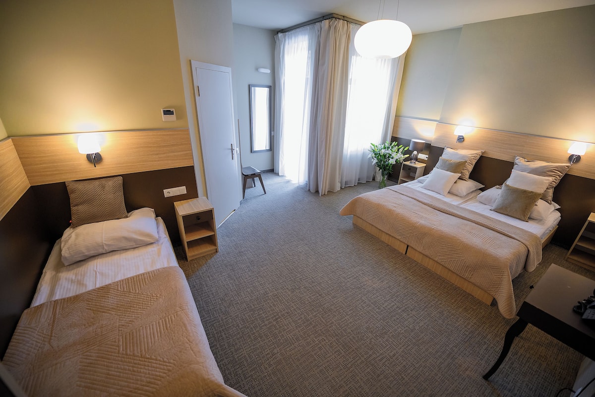Quarto de hotel com duas camas, uma escrivaninha, uma cadeira, uma mesinha e uma planta.