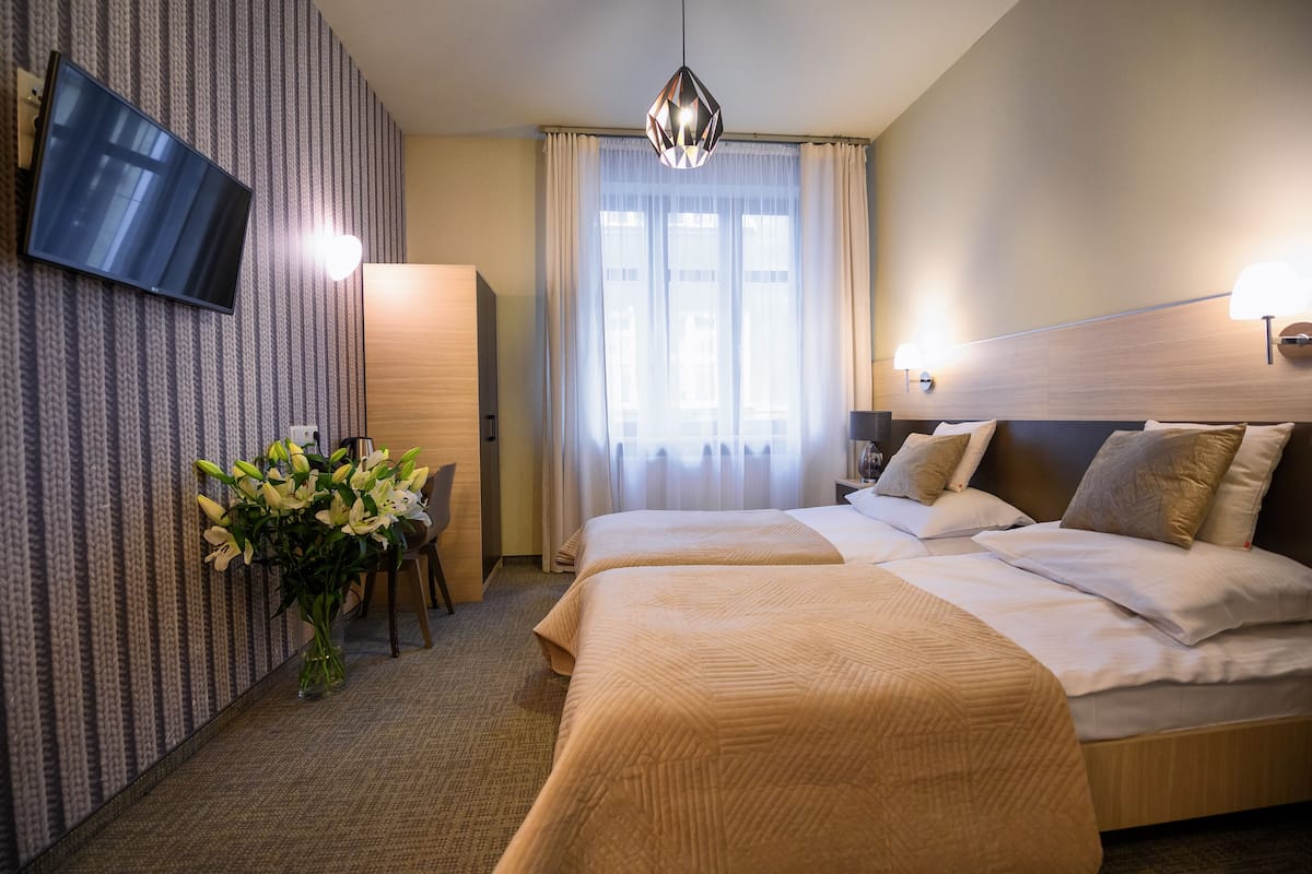 Quarto de hotel com duas camas, televisão presa na parede, escrivaninha com cadeira e janela com cortinas.