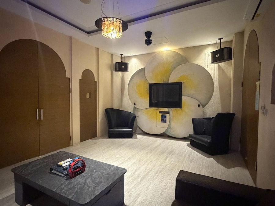 Quarto com cama, duas poltronas, uma mesa de centro, televisão e um lustre.