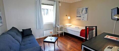 Quarto compacto com sofá, uma pequena mesa, cama com travesseiro vermelho, luminária de cabeceira e telefone.