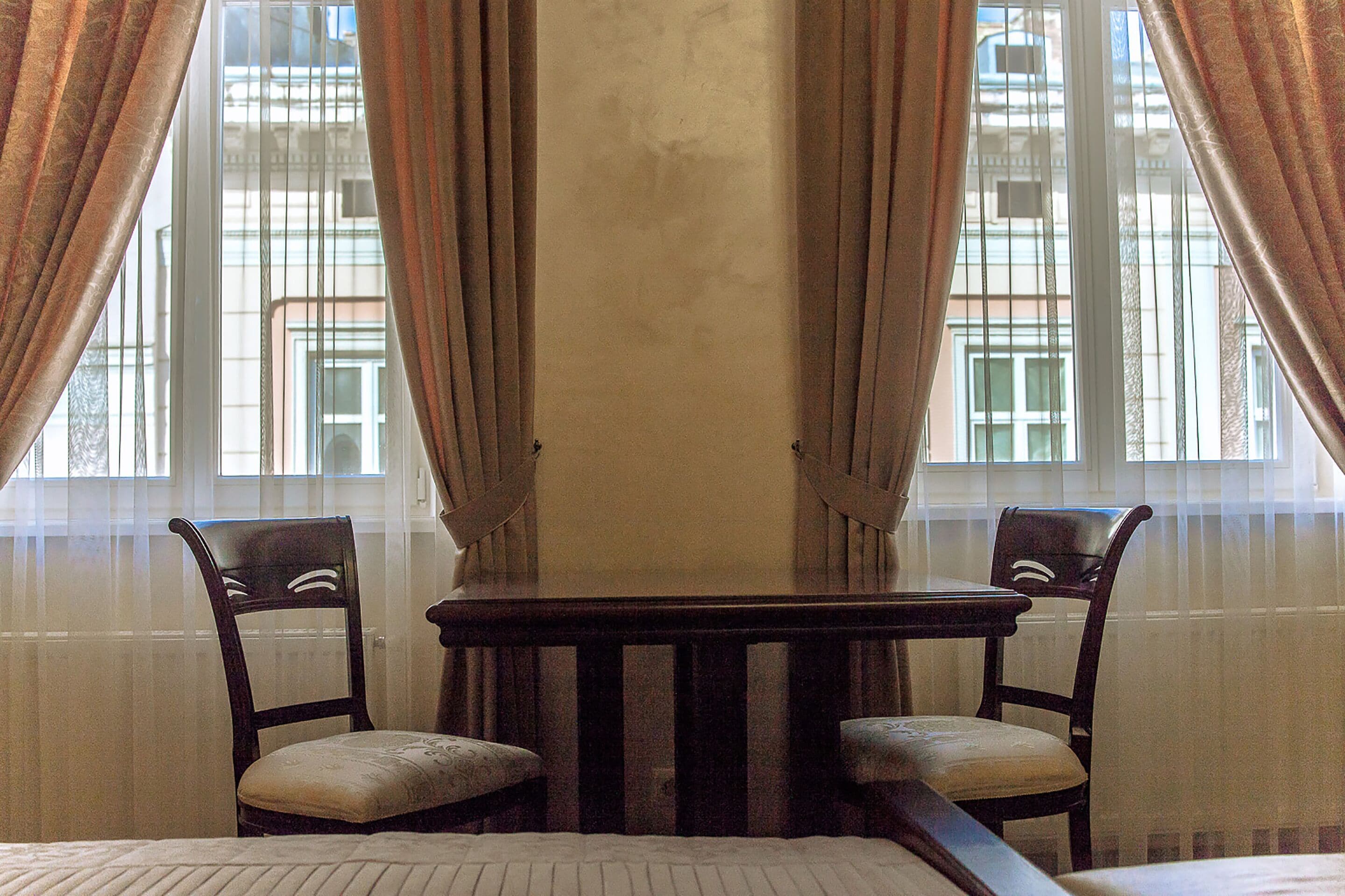 Um quarto com uma cama, duas cadeiras de madeira, uma mesa e uma janela com cortinas.