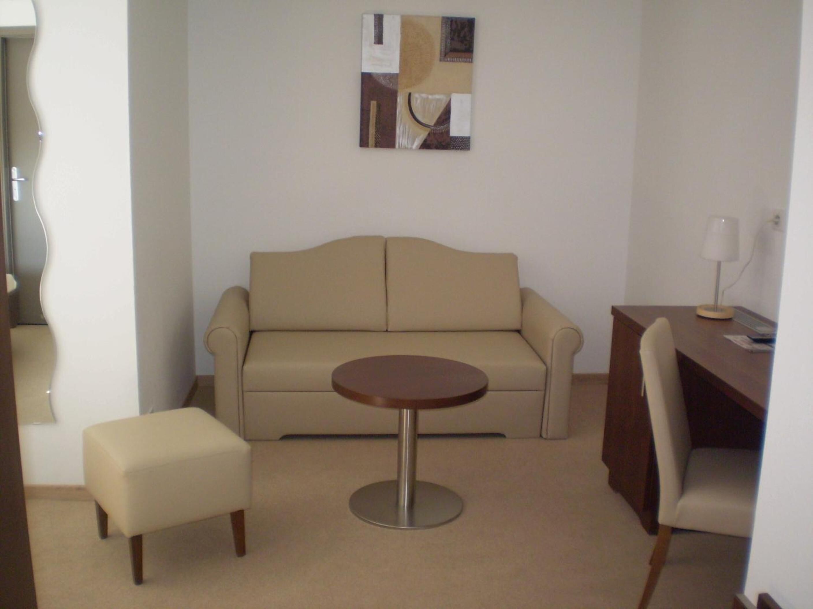 Quarto pequeno e moderno com sofá bege, mesa redonda, um banquinho e escrivaninha com cadeira.