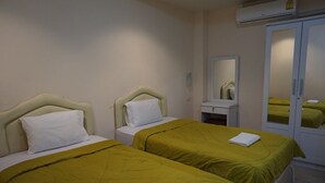 Quarto de hotel com duas camas, cada uma com colcha verde e almofadas brancas. Há um guarda-roupa com espelho e uma pequena mesa com abajur.