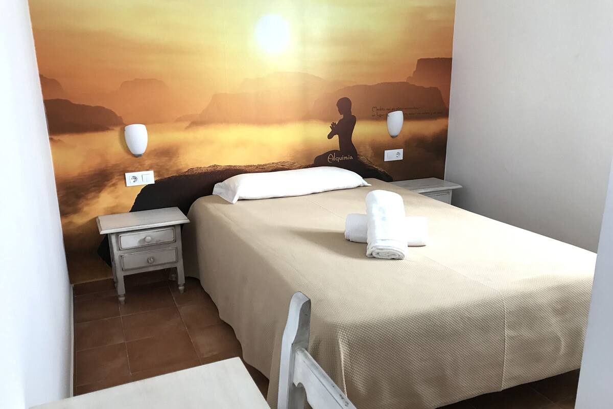 Quarto de hotel com cama, mesa de cabeceira e um mural na parede retratando uma paisagem tranquila.