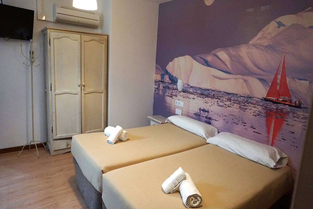 Quarto de hotel com duas camas, um guarda-roupa, televisão de tela plana e um mural de um veleiro numa praia.