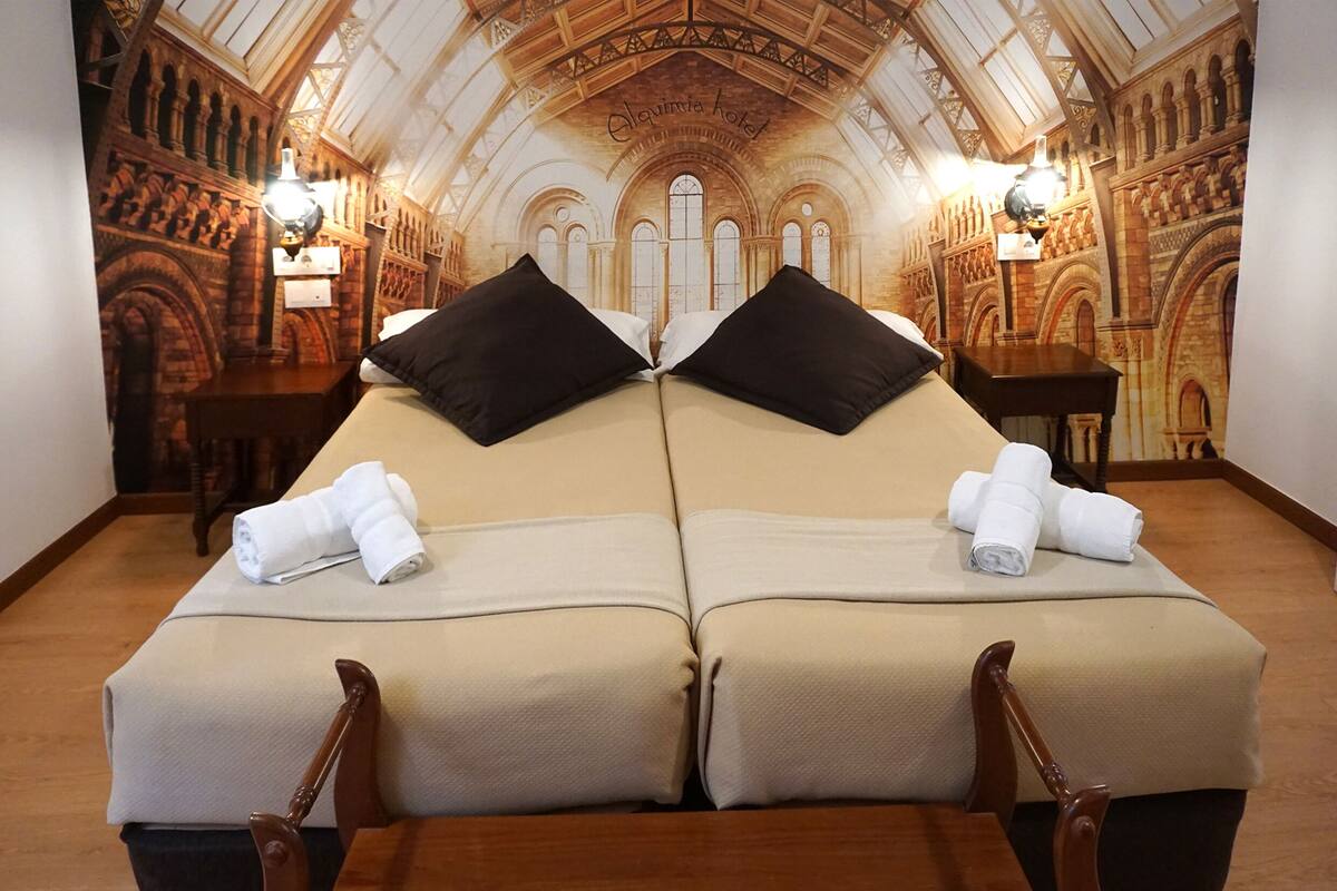 Uma cama de casal com duas almofadas pretas e duas toalhas enroladas, num quarto com teto abobadado e janelas em arco.