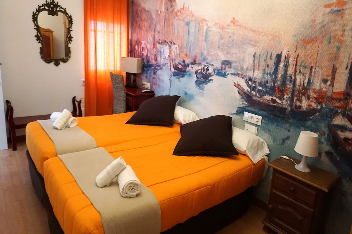 Quarto de hotel com duas camas, uma pintura grande de uma paisagem urbana, um espelho e uma mesa de cabeceira com abajur.