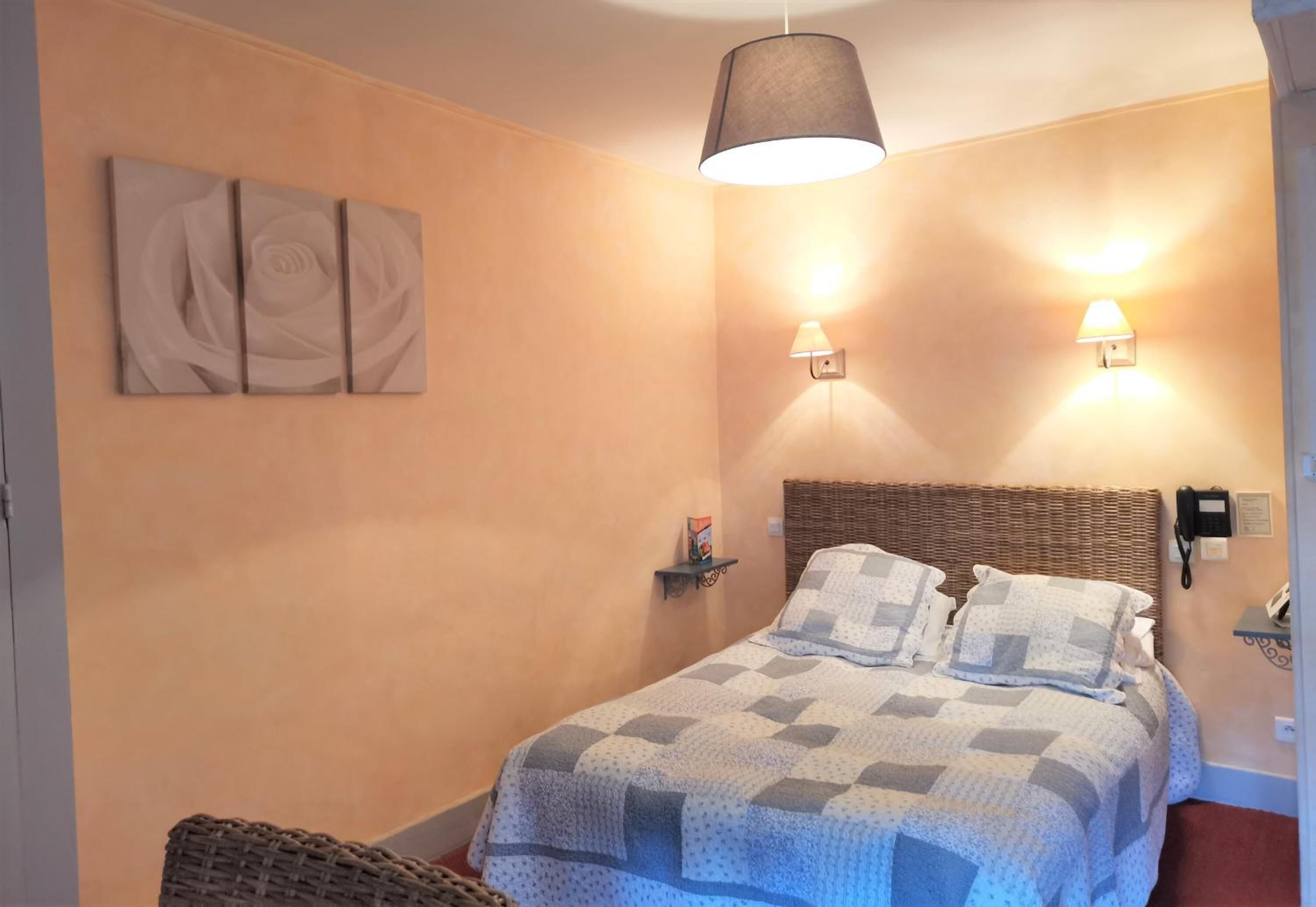 Quarto com cama, duas luminárias de parede, um candeeiro pendente, uma obra de arte na parede e uma estante com uma caixa.