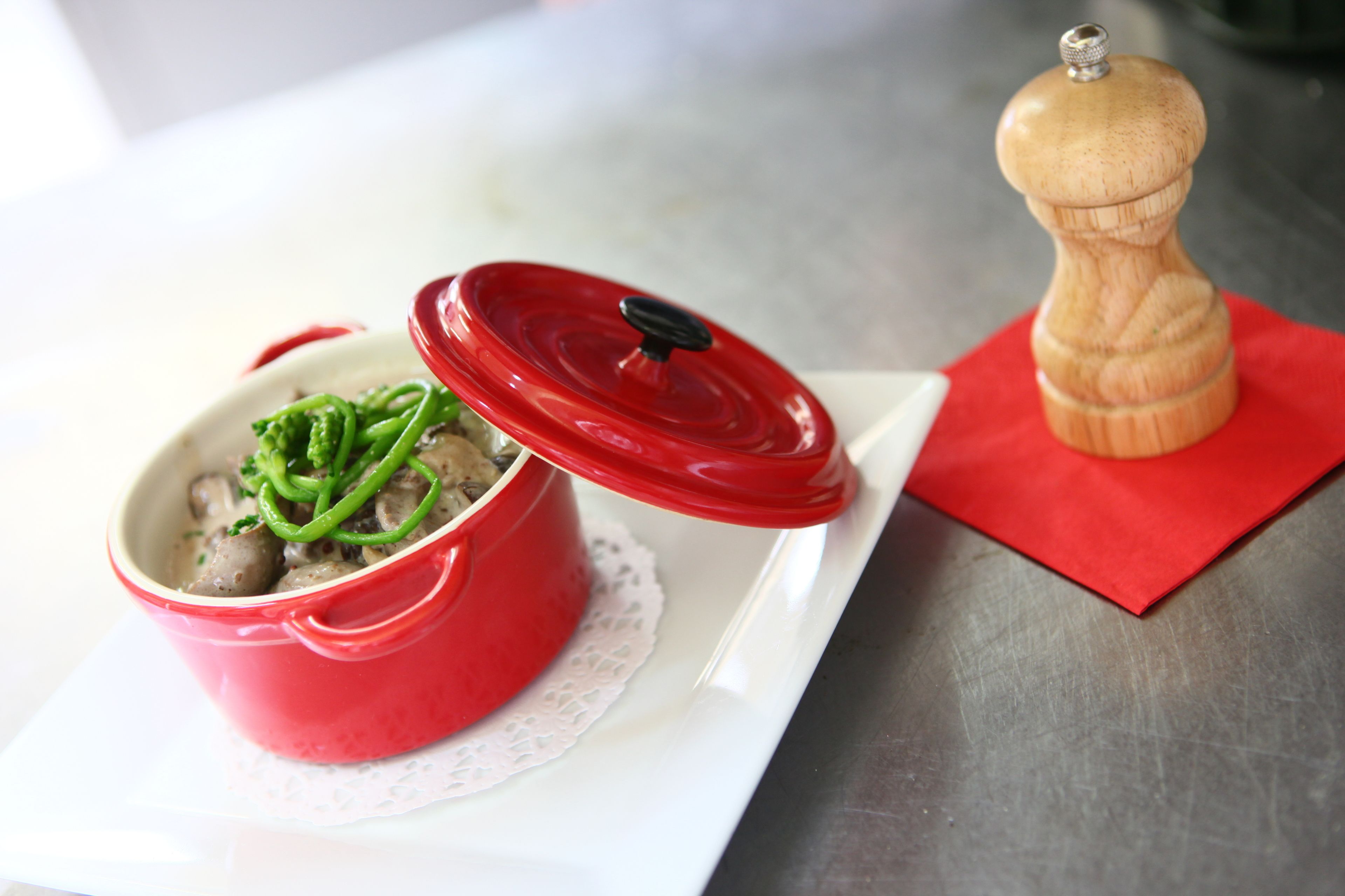 Um pote vermelho com tampa sobre um prato branco, um moedor de pimenta de madeira e um guardanapo vermelho.