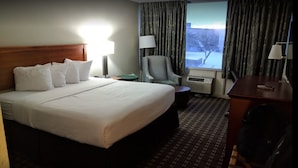 Quarto de hotel com uma cama grande, escrivaninha, cadeira e janela com cortinas.