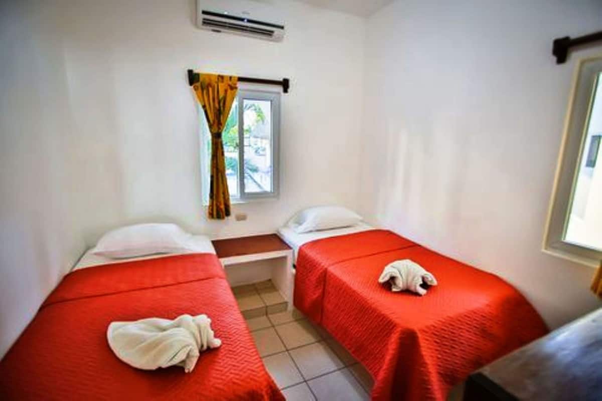 Quarto pequeno e limpo com duas camas de solteiro, janela com cortinas e uma mesa.