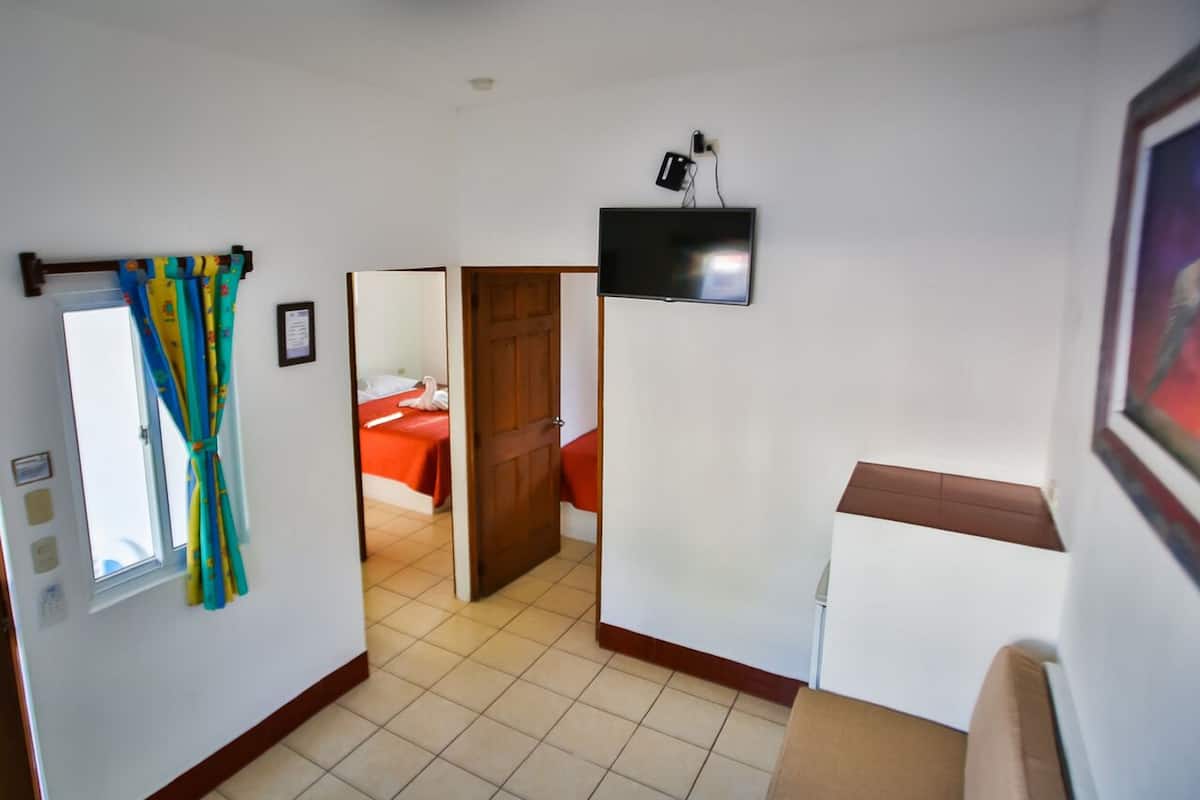 Quarto de hotel com cama, televisão fixada na parede, janela com cortinas e porta que dá para outro cômodo.