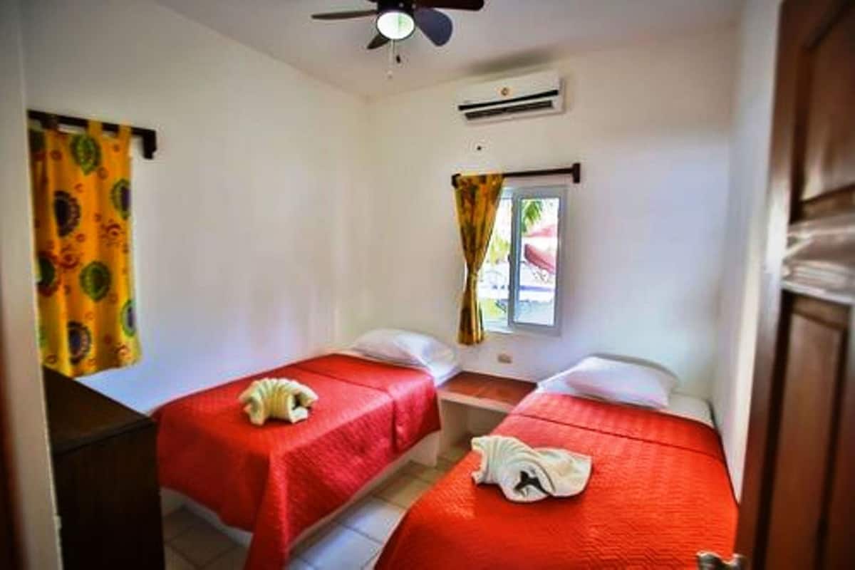Quarto de hotel com duas camas de solteiro, ventilador de teto, ar condicionado e janela com cortinas.