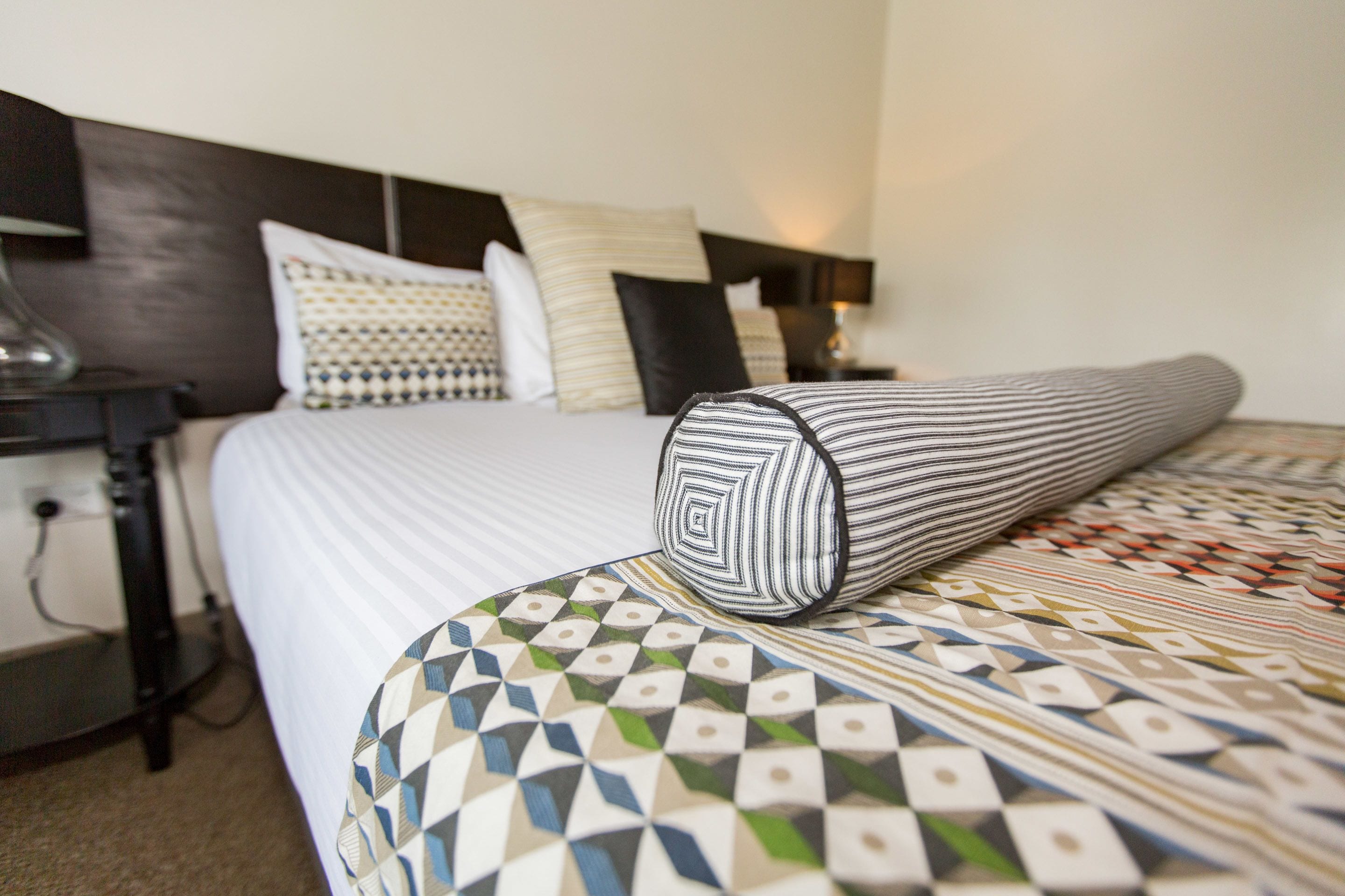Uma cama bem arrumada com roupa de cama estampada, uma mesa de cabeceira com abajur e um criado-mudo fixado na parede.