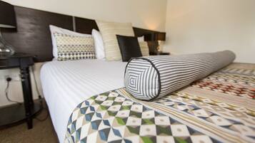Uma cama bem arrumada com roupa de cama estampada, uma mesa de cabeceira com abajur e um criado-mudo fixado na parede.