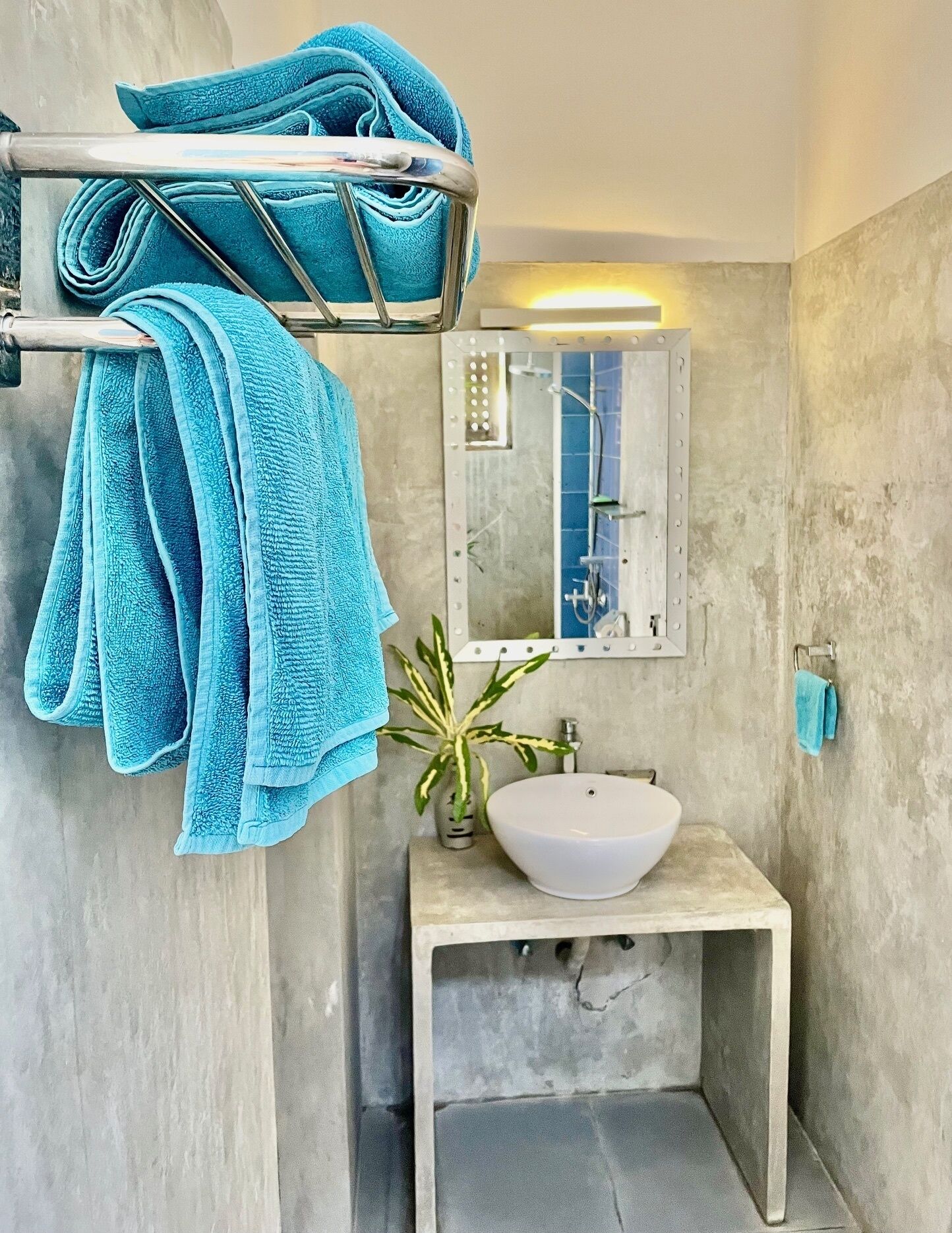Casa de banho com parede de concreto, lavatório branco, espelho e porta-toalhas com toalhas azuis.