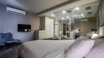 Quarto de hotel com uma cama grande, televisão de tela plana, uma cadeira e área de chuveiro com box de vidro.