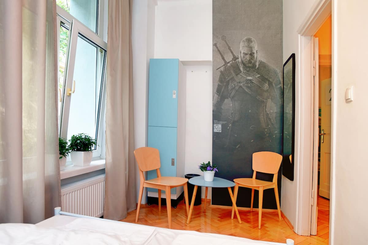 Quarto de hotel com cama, janela com cortinas, radiador, armário azul, duas cadeiras de madeira, uma mesinha com uma planta e um pôster grande na parede.