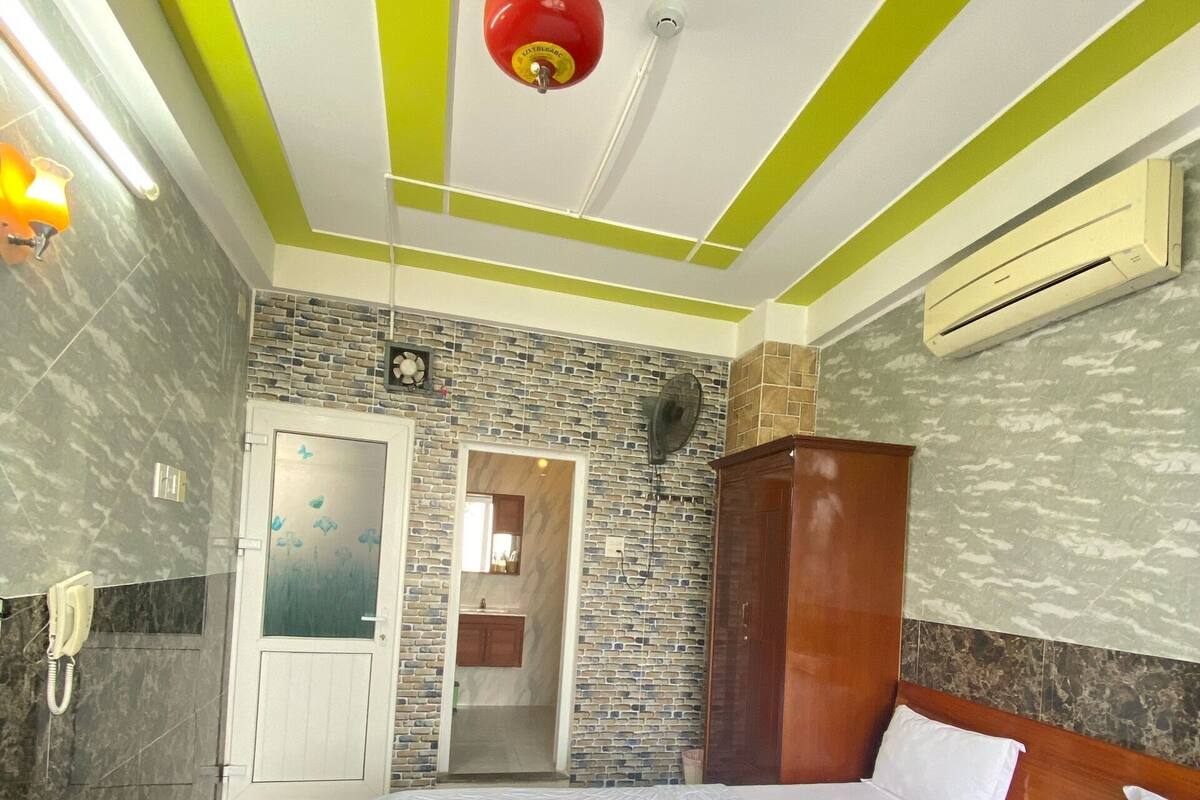 Quarto de hotel com cama, guarda-roupa, banheiro com espelho e alarme de incêndio vermelho.