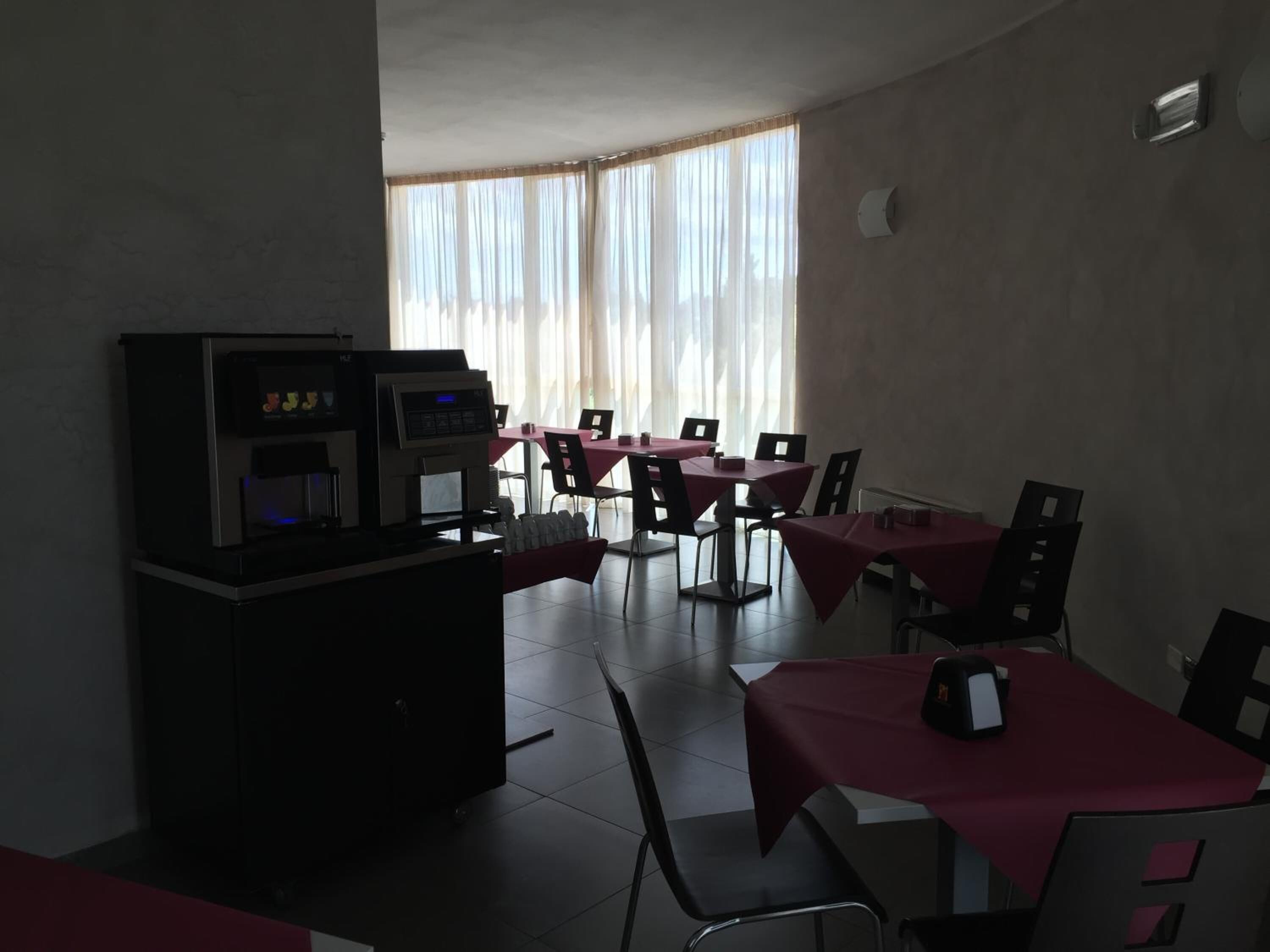 Uma área de refeições com mesas cobertas por toalhas vermelhas, uma máquina de café e cadeiras.