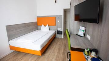 Quarto de hotel com uma cama de solteiro, televisão fixada na parede, mesa de cabeceira pequena e uma escrivaninha com cadeira.