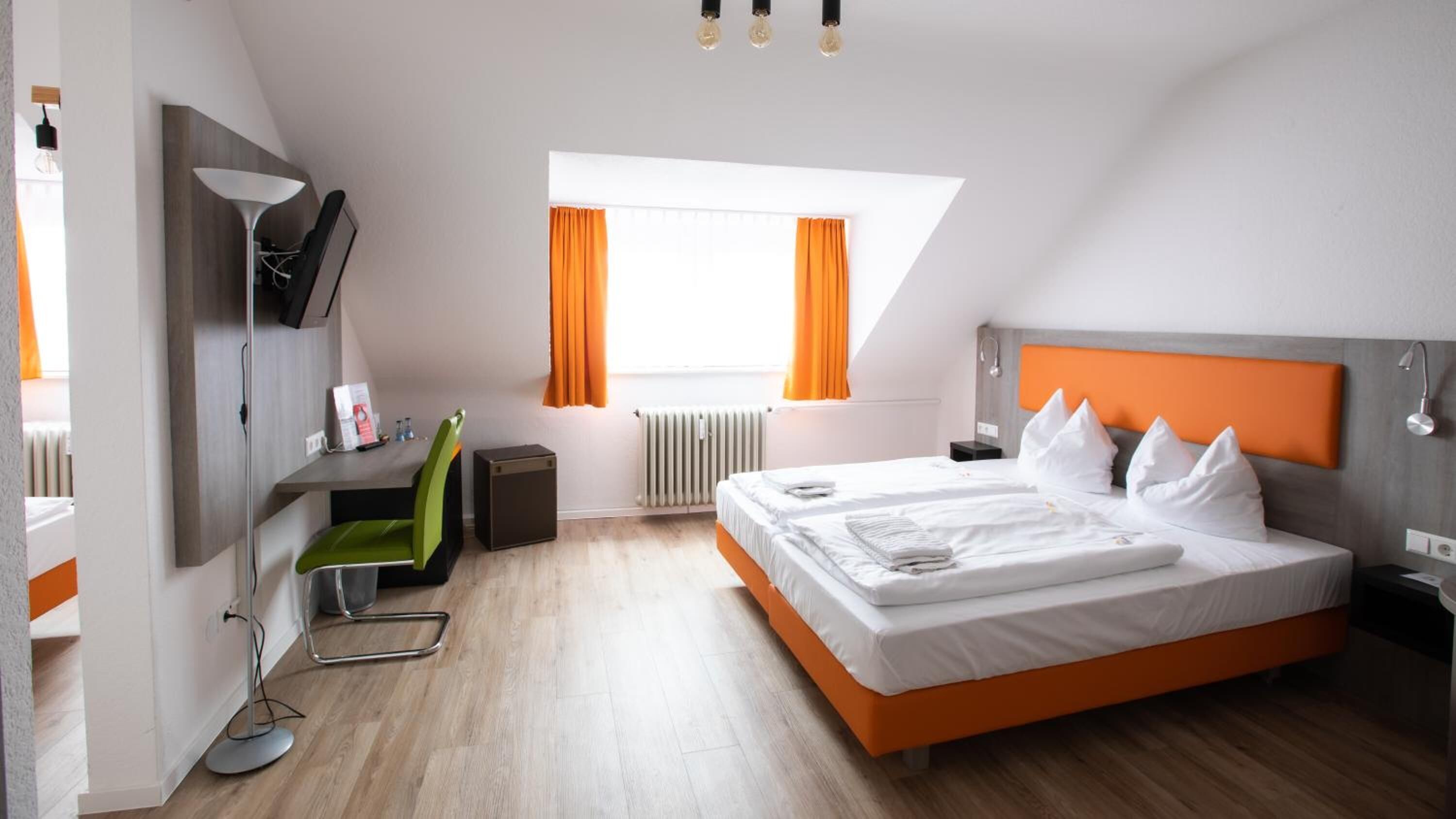Quarto de hotel moderno com uma cama grande, uma escrivaninha, uma cadeira, um radiador e uma janela com cortinas.