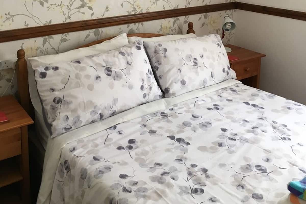 Uma cama com roupa de cama floral, cabeceira de madeira, duas mesas de cabeceira e uma mesa de apoio com luminária.