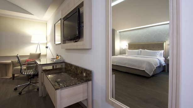 Quarto de hotel com uma cama, uma escrivaninha com cadeira, uma televisão fixada na parede e um espelho refletindo o aposento.