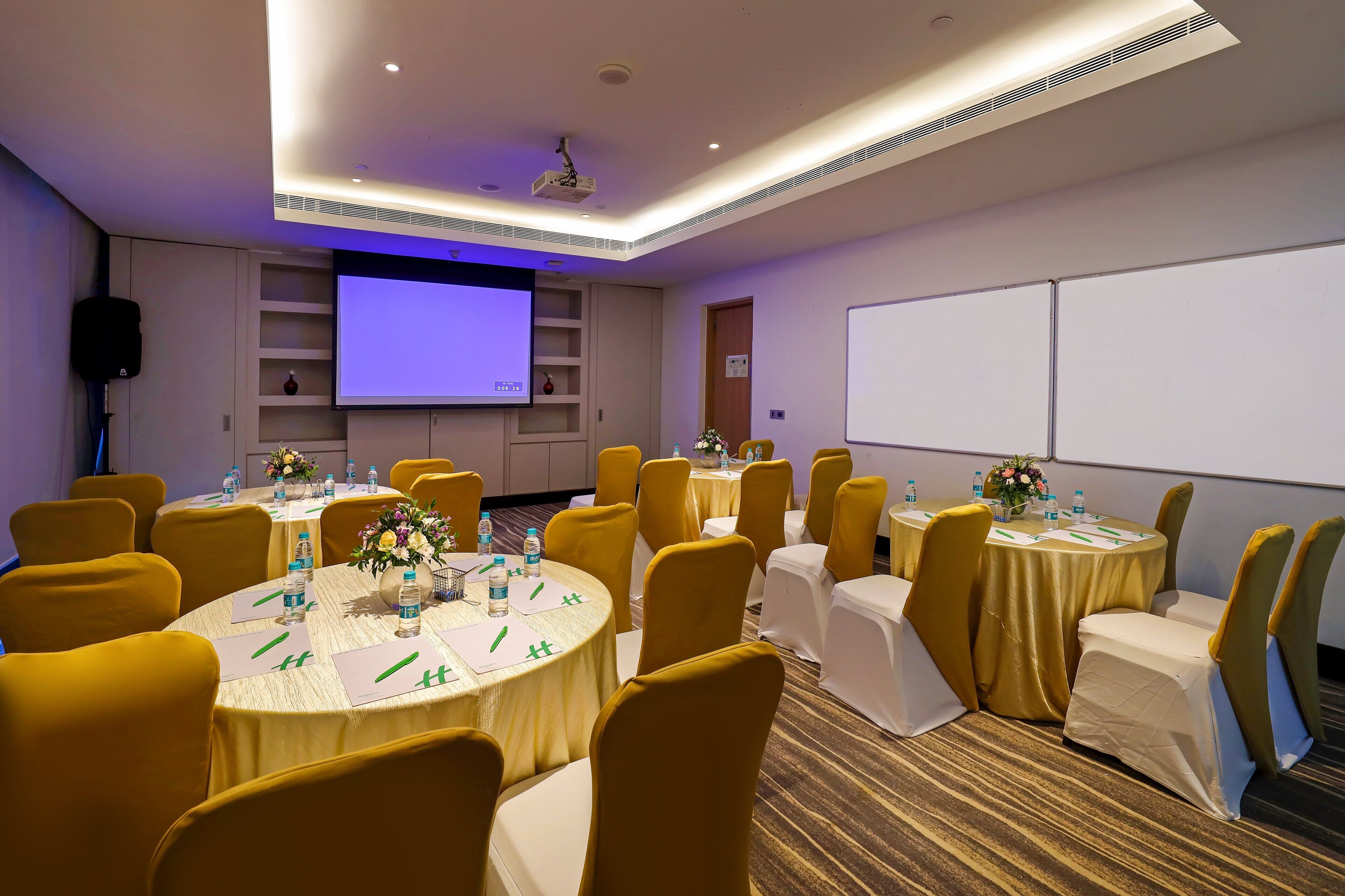 Uma sala de conferências com várias mesas, cadeiras amarelas e uma tela de projeção.