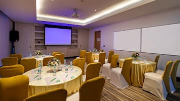 Uma sala de conferências com várias mesas, cadeiras amarelas e uma tela de projeção.