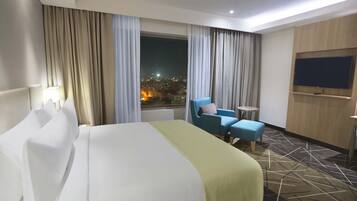 Quarto de hotel com uma cama grande, uma poltrona azul, uma mesa pequena e uma televisão fixada na parede.