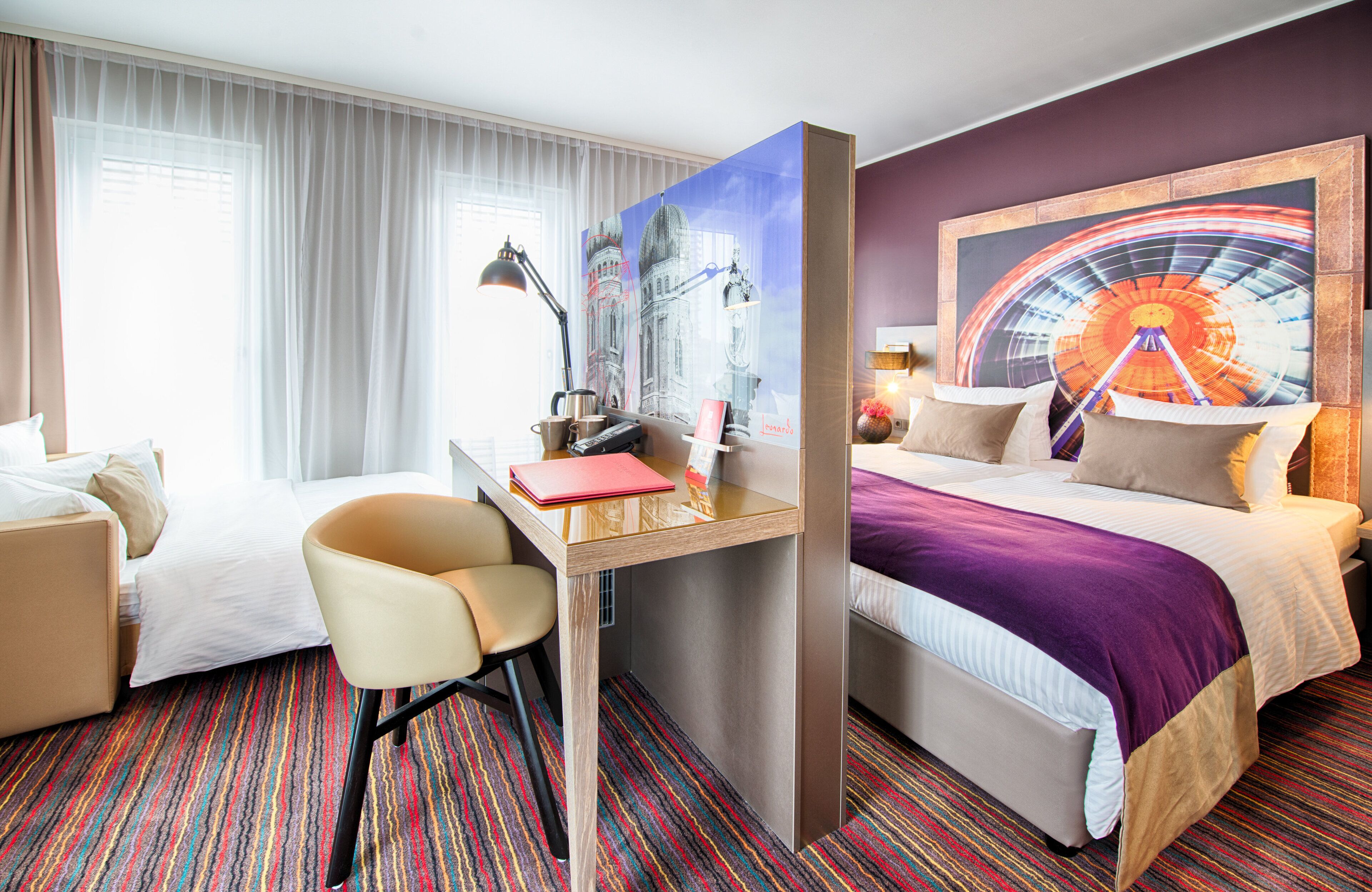 Quarto de hotel com uma cama, uma escrivaninha com cadeira, uma janela com cortinas e um grande quadro na parede.