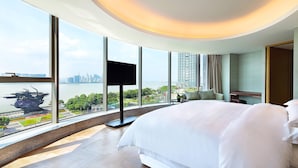 Quarto de hotel moderno com uma cama grande, televisão e vista para a paisagem urbana e rio.