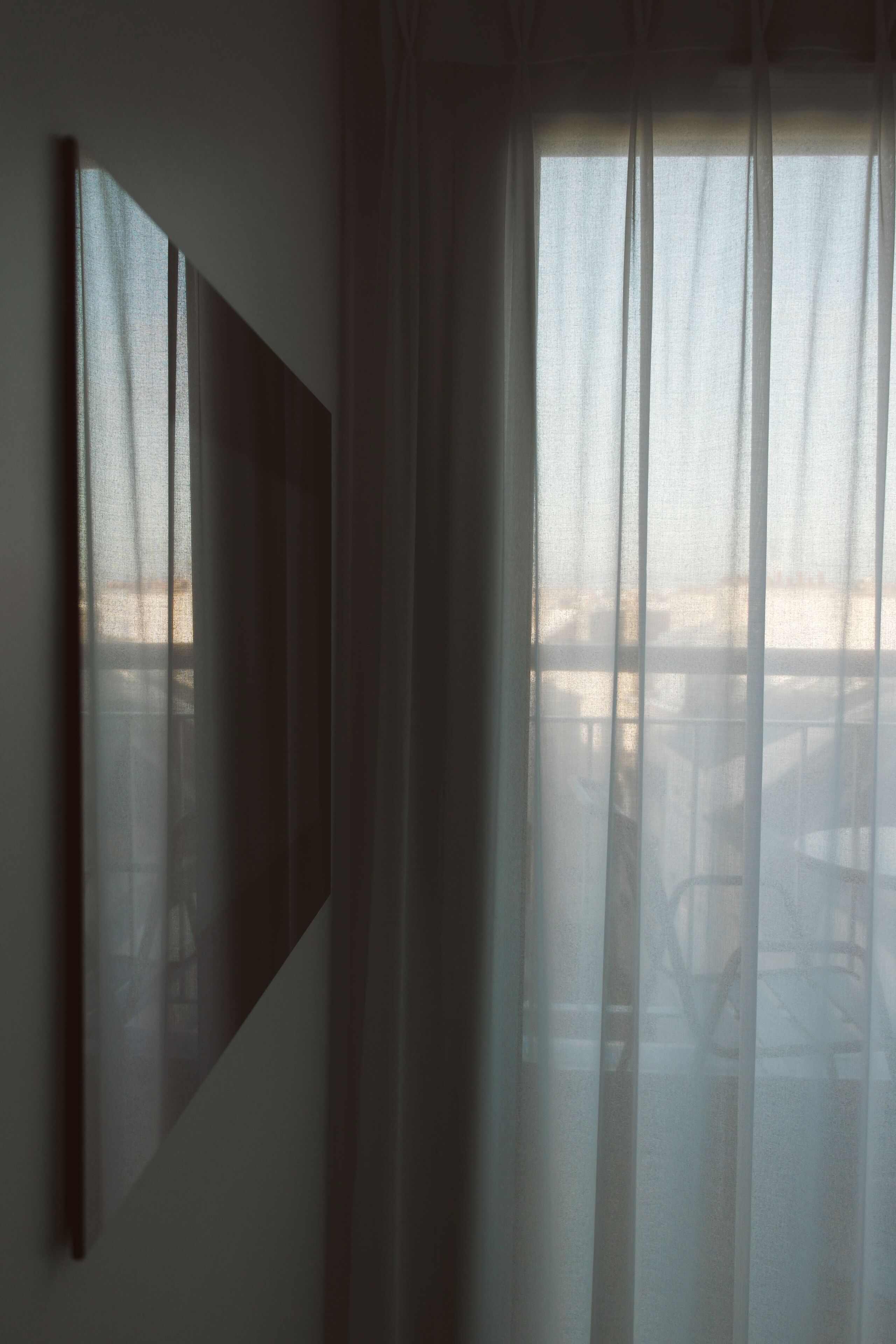 Quarto com uma grande janela coberta por cortinas leves, um quadro na parede e um vislumbre de móveis de exterior.