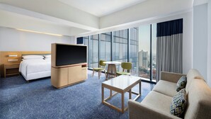 Quarto de hotel com cama, televisão, sofá, mesa e vista para a cidade através de amplas janelas.