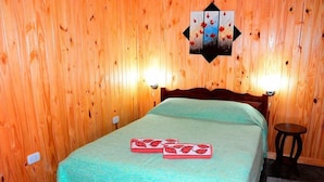 Quarto com painéis de madeira, uma cama, duas luminárias fixadas na parede, um banquinho pequeno e uma obra de arte na parede.
