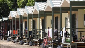 Uma fileira de pequenas cabanas térreas, com telhados de metal e pilares verdes, cada uma com uma pequena varanda.