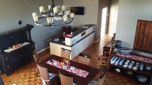 Uma sala de jantar com uma mesa de madeira, cadeiras, um lustre e uma cozinha ao fundo.
