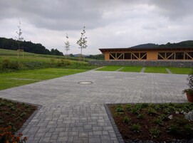 Um edifício com fachada de pedra e grandes janelas, rodeado por um pátio pavimentado com uma pequena área de jardim.
