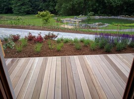 Um deck de madeira com vista para um jardim paisagístico, com diversas plantas e um pequeno lago.