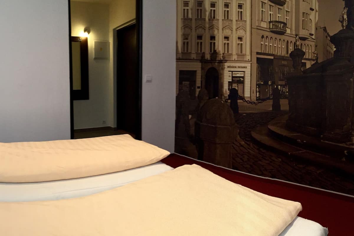 Quarto de hotel com duas camas, um espelho grande e vista para uma rua da cidade com edifícios e uma fonte.
